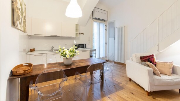 Cozinha moderna com armários brancos, uma mesa de jantar de madeira e um sofá branco numa sala de estar iluminada.
