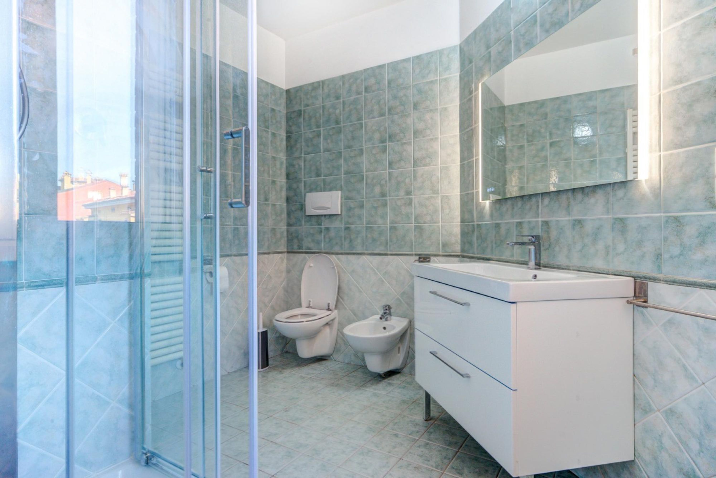 Casa de banho com cabine de duche em vidro, sanita, bidé e toucador branco com espelho.