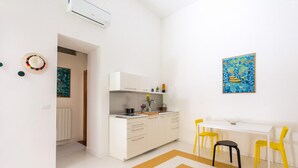 Uma cozinha compacta com armários brancos, bancada branca e uma pequena área de refeições com cadeiras amarelas.
