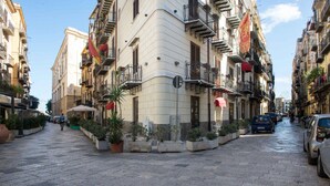 Uma rua de paralelepípedos estreita, ladeada por edifícios altos, com varandas adornadas com plantas e bandeiras, e carros estacionados em ambos os lados.