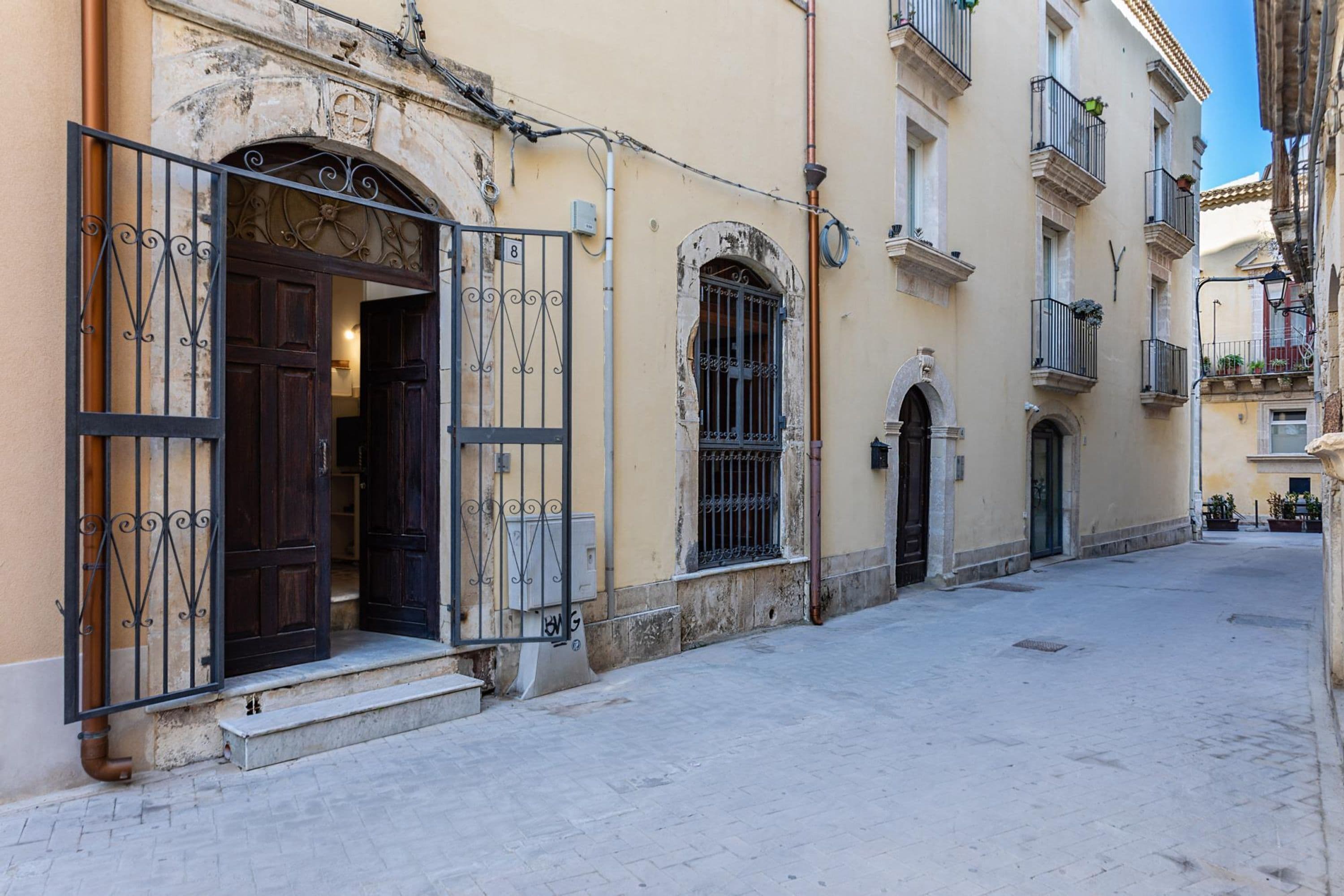 Uma rua estreita com um edifício que apresenta um portão aberto e uma porta de madeira, uma janela com grade de metal e uma pequena varanda com vasos de plantas.