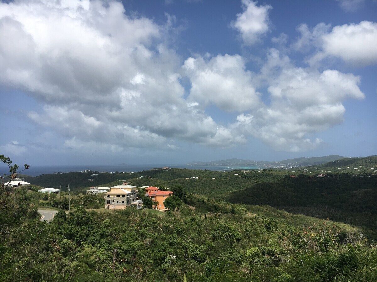 Paisagem costeira com edifícios numa encosta, estrada sinuosa e vista para o mar e colinas ao longe.