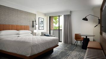 Quarto de hotel com uma cama grande, uma escrivaninha, uma cadeira, uma mesinha e uma janela com cortinas.