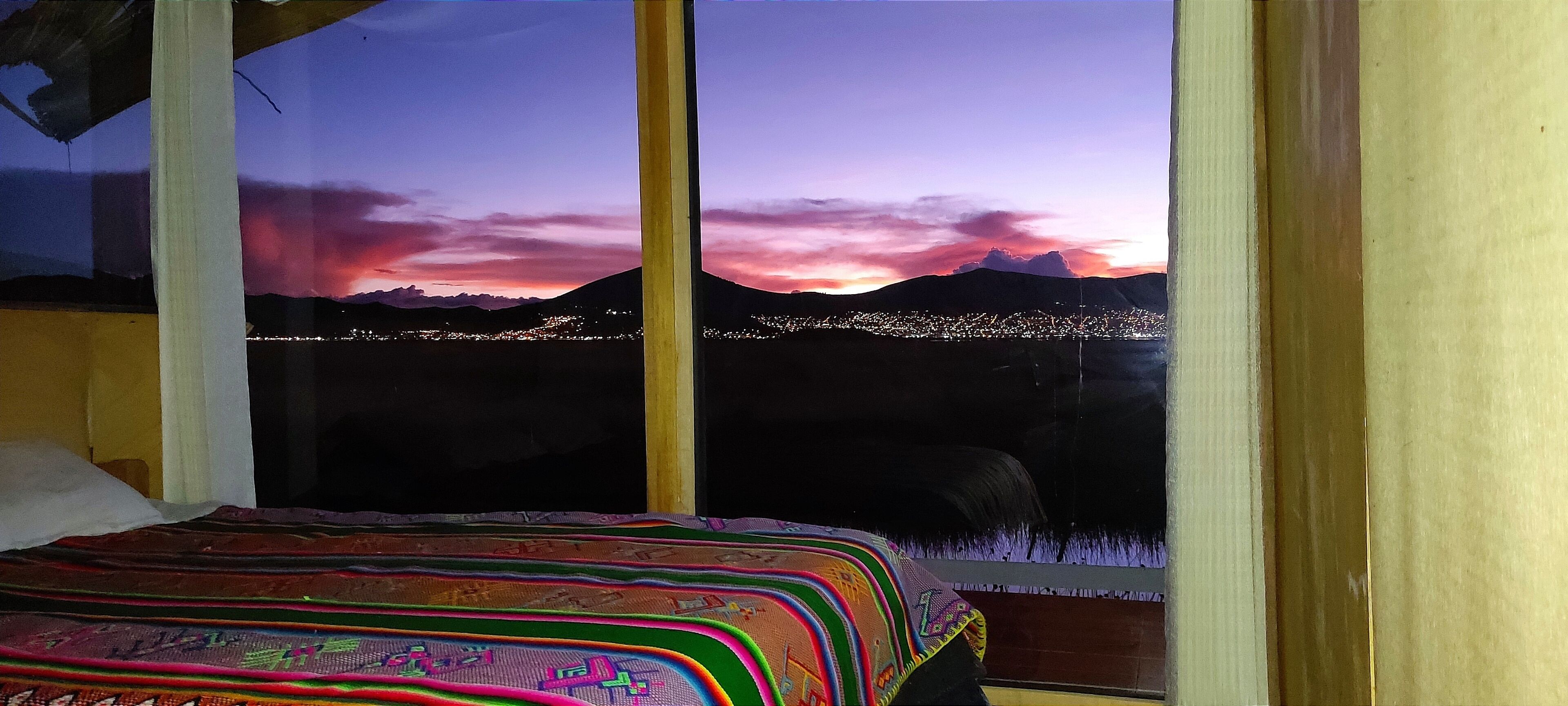 Quarto com cama, uma janela grande e vista para a cidade ao entardecer.