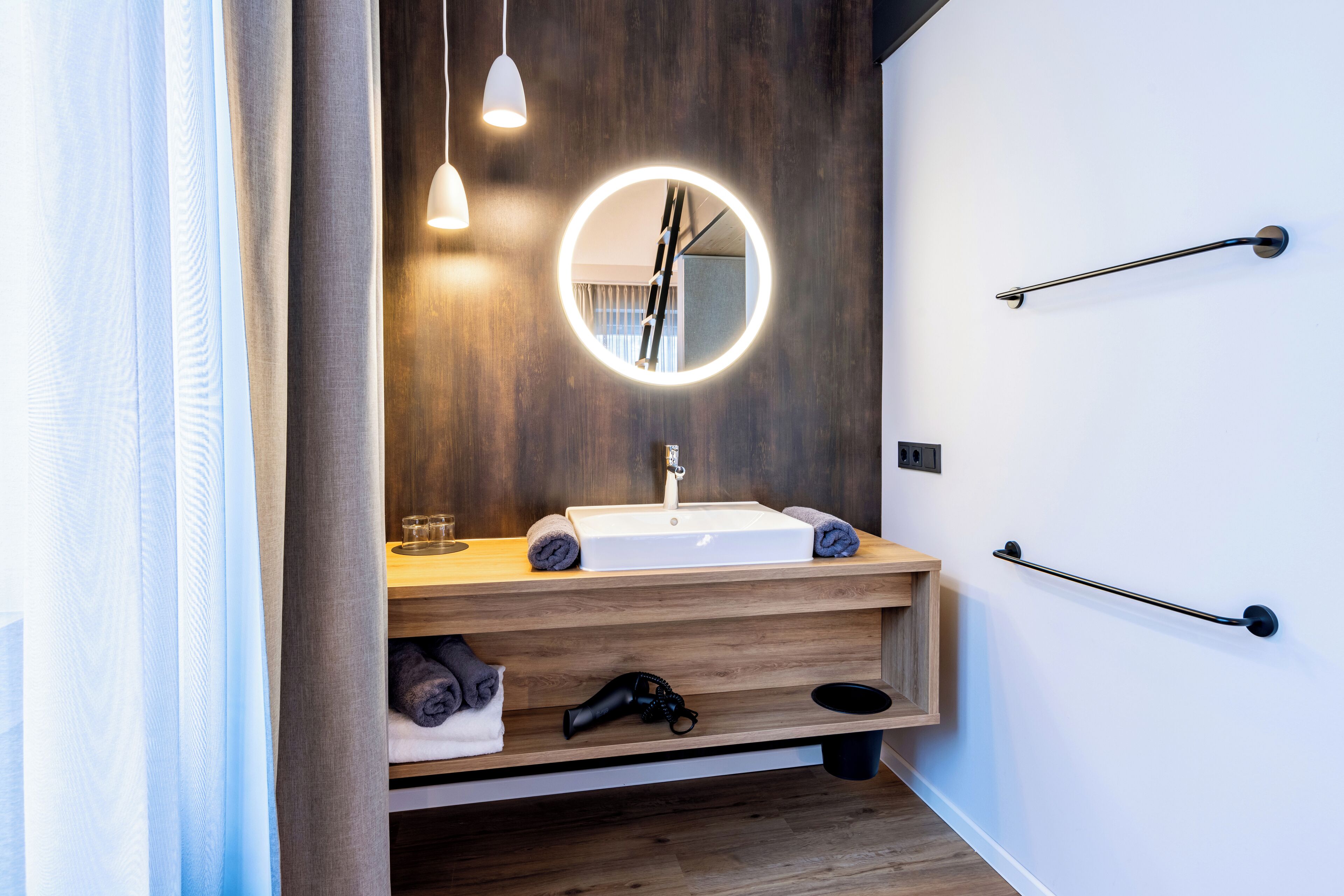Uma casa de banho moderna com uma toucador de madeira, um espelho redondo com luz e um porta-toalhas.