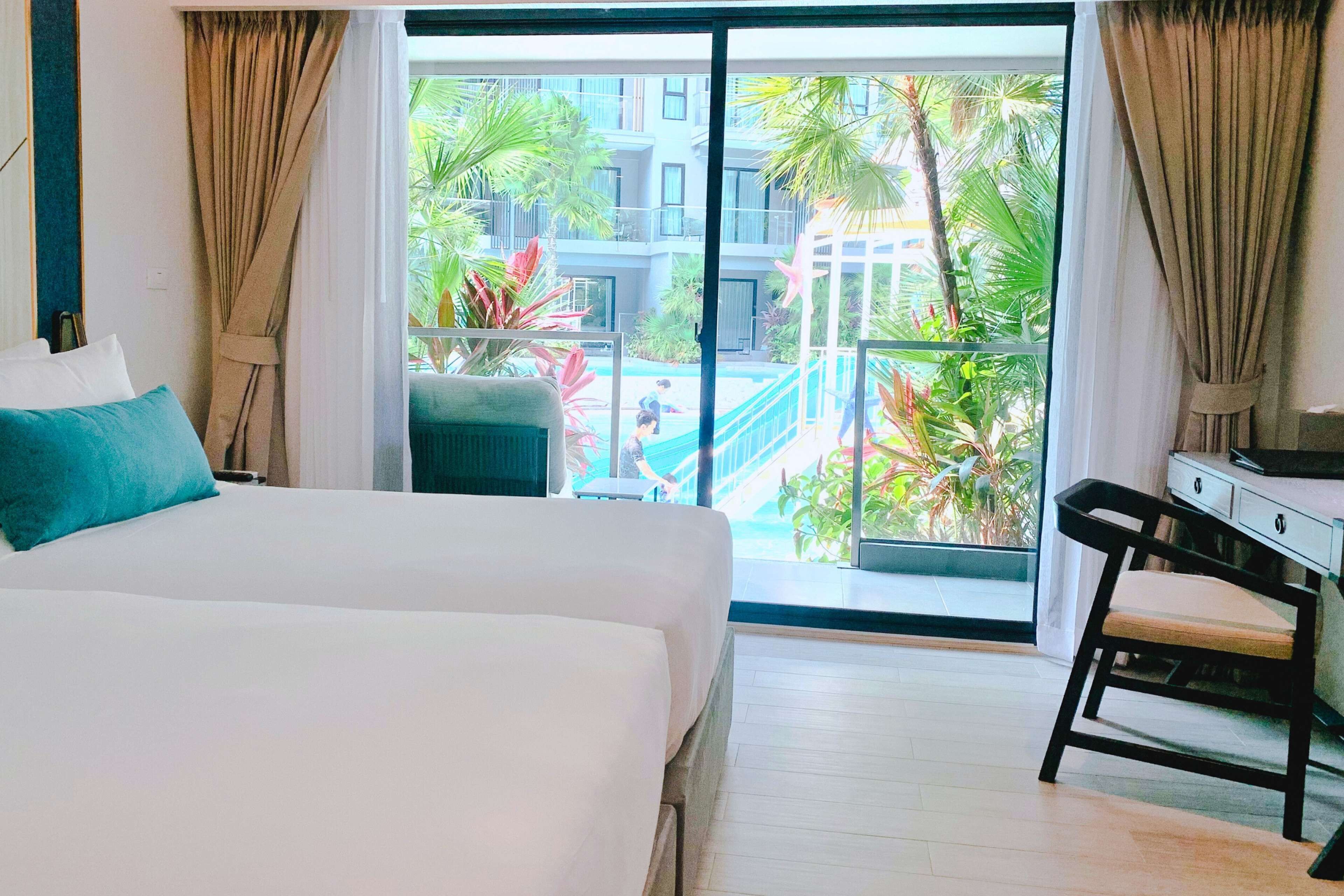 Quarto de hotel com uma cama grande, uma escrivaninha e vista para uma piscina e vegetação.