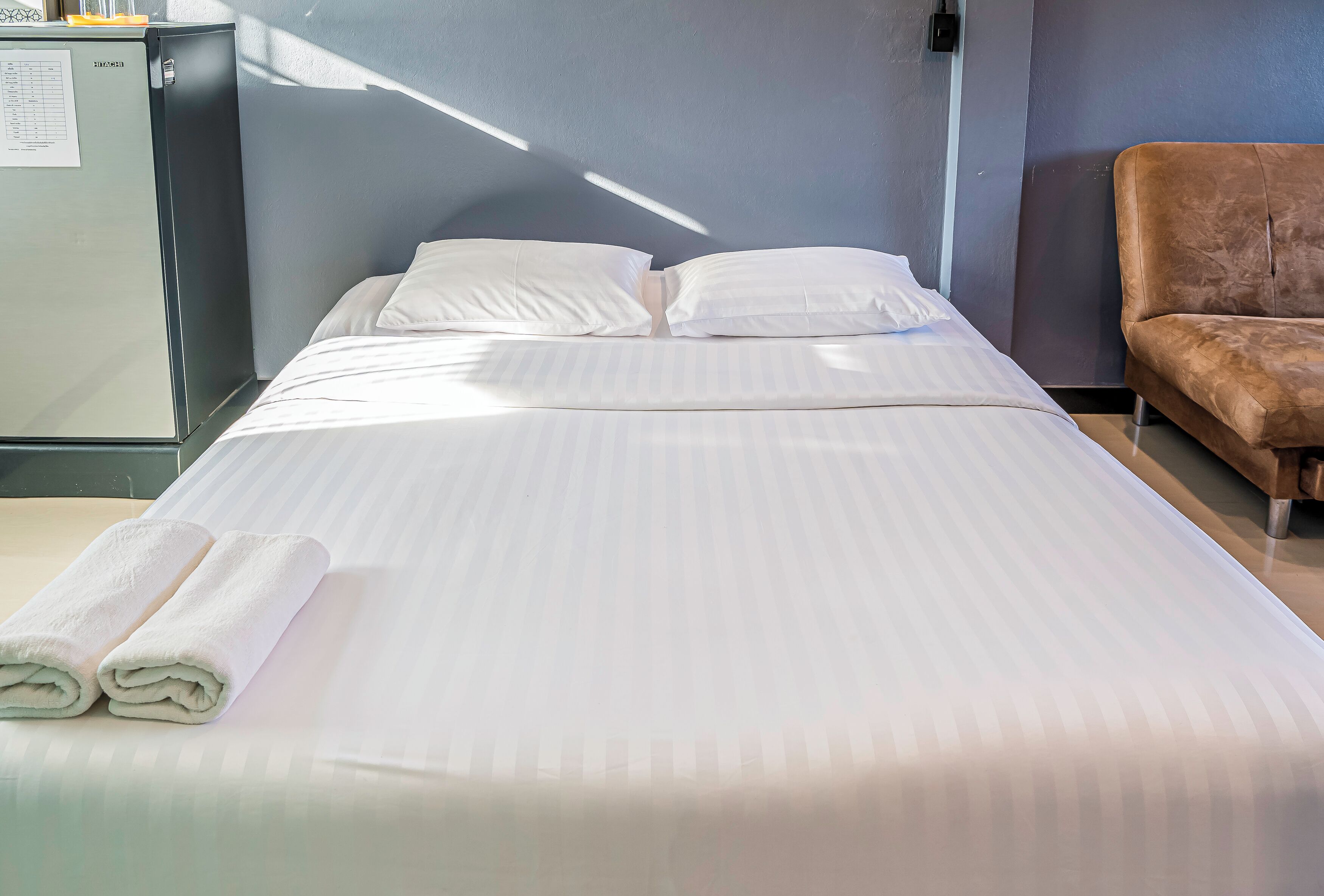 Uma cama bem arrumada com lençóis brancos e duas toalhas enroladas, uma poltrona estofada marrom e uma pequena cozinha compacta com geladeira.