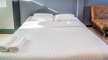 Uma cama bem arrumada com lençóis brancos e duas toalhas enroladas, uma poltrona estofada marrom e uma pequena cozinha compacta com geladeira.