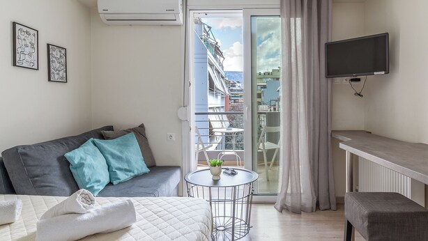 Quarto de hotel moderno com varanda, sofá, uma pequena mesa com uma planta e televisão fixada na parede.