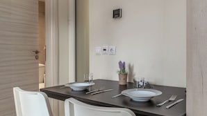 Uma área de refeições com uma mesa escura posta para dois, cadeiras brancas, um vaso de lavanda e um pequeno dispositivo fixado na parede.