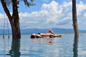 Uma pessoa deitada em uma plataforma flutuante em um corpo d'água, com árvores e montanhas ao fundo.