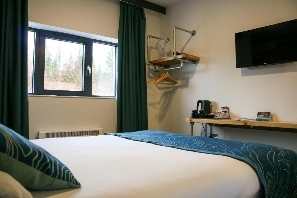 Quarto de hotel com cama, televisão presa na parede, janela com cortinas e uma pequena mesa com uma cafeteira e alguns objetos.