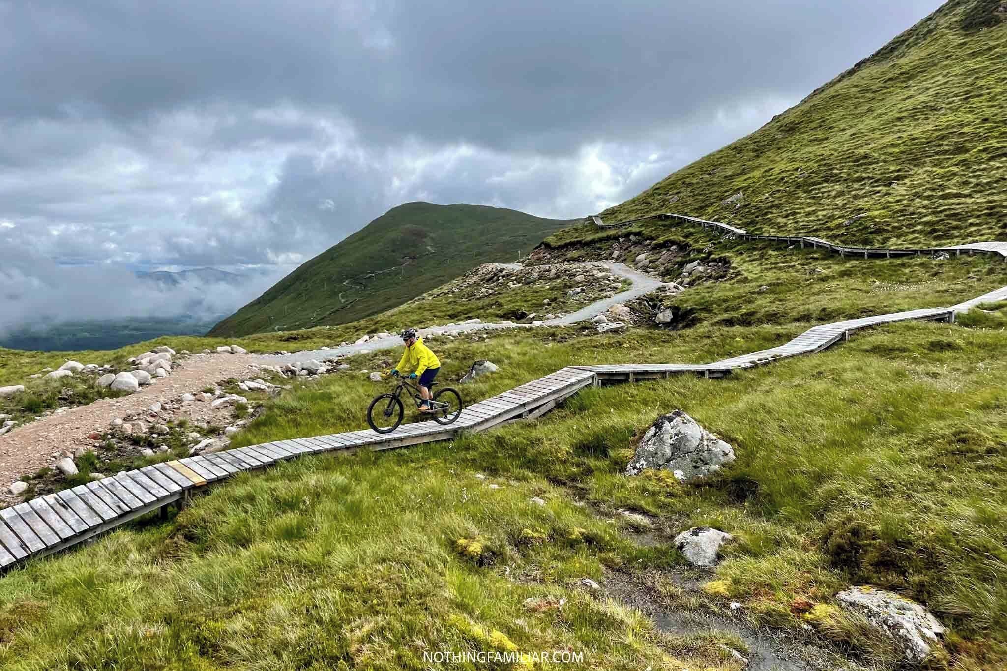 Uma trilha na montanha com pontes de madeira e um ciclista usando jaqueta amarela.