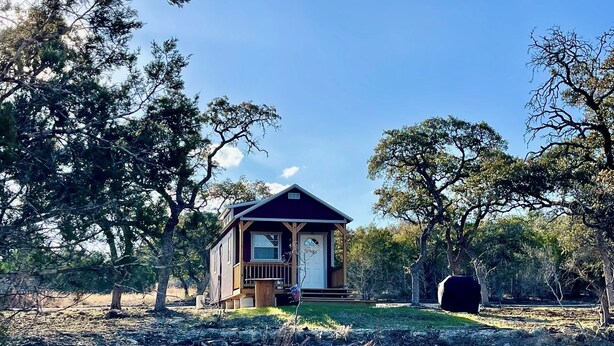 Uma cabana pequena e térrea com varanda, rodeada por árvores e uma área gramada.