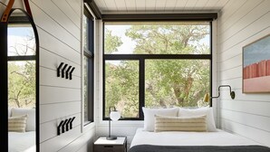 Quarto com uma janela grande, mesa de cabeceira com abajur e cama com travesseiros.
