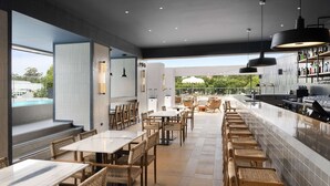 Uma área de refeições externa moderna com mesas e cadeiras de madeira, um bar com painel de azulejos e vista para a piscina.
