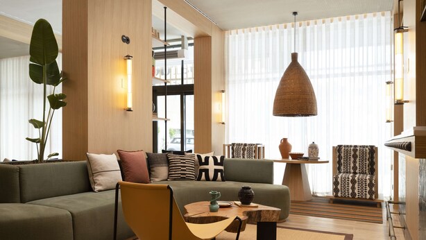 Uma sala de estar moderna com sofá, poltrona e mesa de centro, com uma planta grande, iluminação pendente e vista através de janelas amplas.