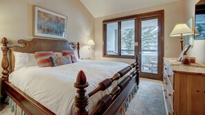 Quarto com uma cama grande, cabeceira de madeira, abajures, cômoda e janela com vista para árvores.