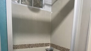 Um chuveiro pequeno e fechado, com um chuveiro fixado na parede, uma prateleira de metal para pendurar objetos e piso revestido de azulejos.