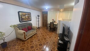 Uma sala de estar com piso estampado, um sofá, uma mesa de jantar com cadeiras, uma televisão e um vaso com planta.