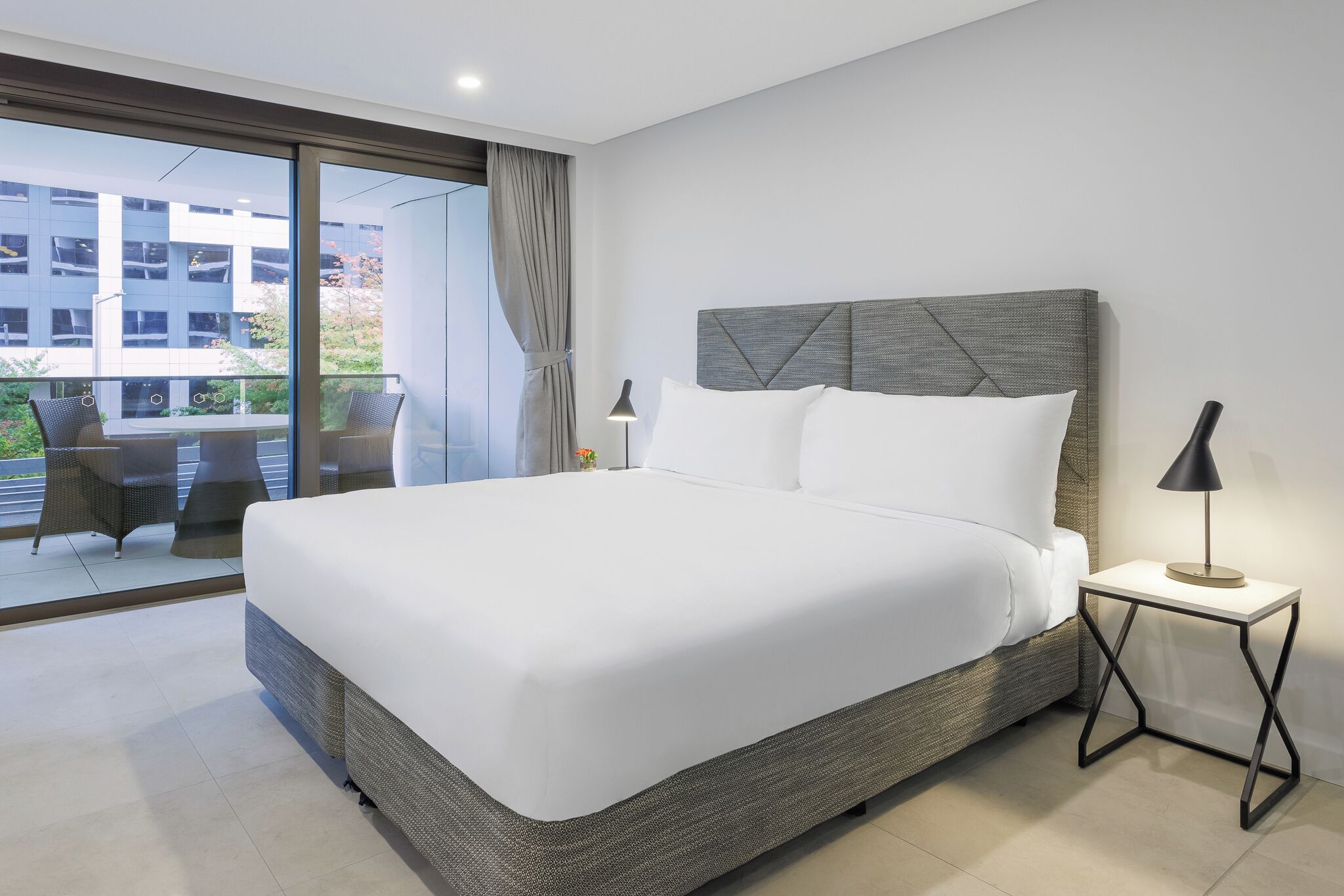 Quarto de hotel moderno com uma cama grande, uma mesinha de cabeceira com abajur e vista para a varanda.