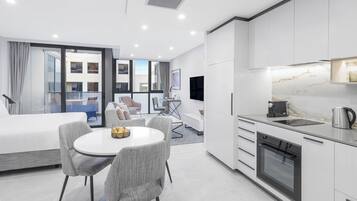 Quarto de hotel moderno com área de refeições, cozinha compacta e uma grande janela com vista para a cidade.