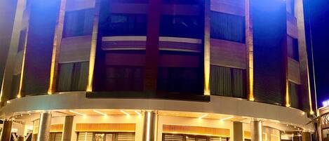 Um hotel moderno com fachada curva, sinalização iluminada e área de entrada bem iluminada com plantas em vasos.