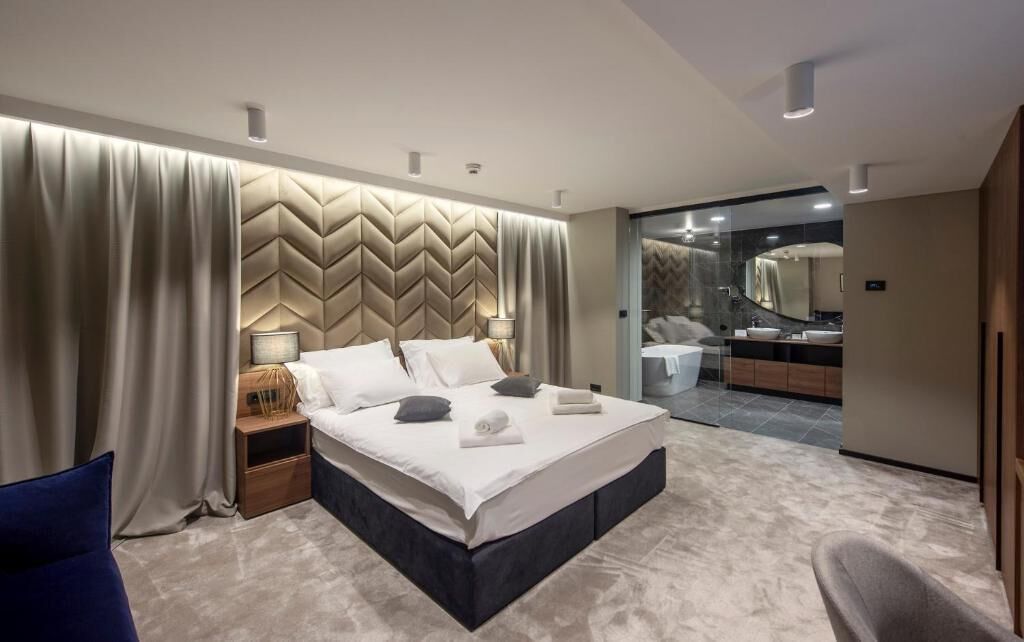 Um quarto moderno com uma cama grande, mesas de cabeceira, um criado-mudo, um espelho e um banheiro visível através de uma porta de vidro.