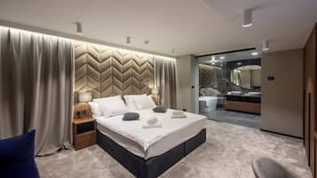 Um quarto moderno com uma cama grande, mesas de cabeceira, um criado-mudo, um espelho e um banheiro visível através de uma porta de vidro.