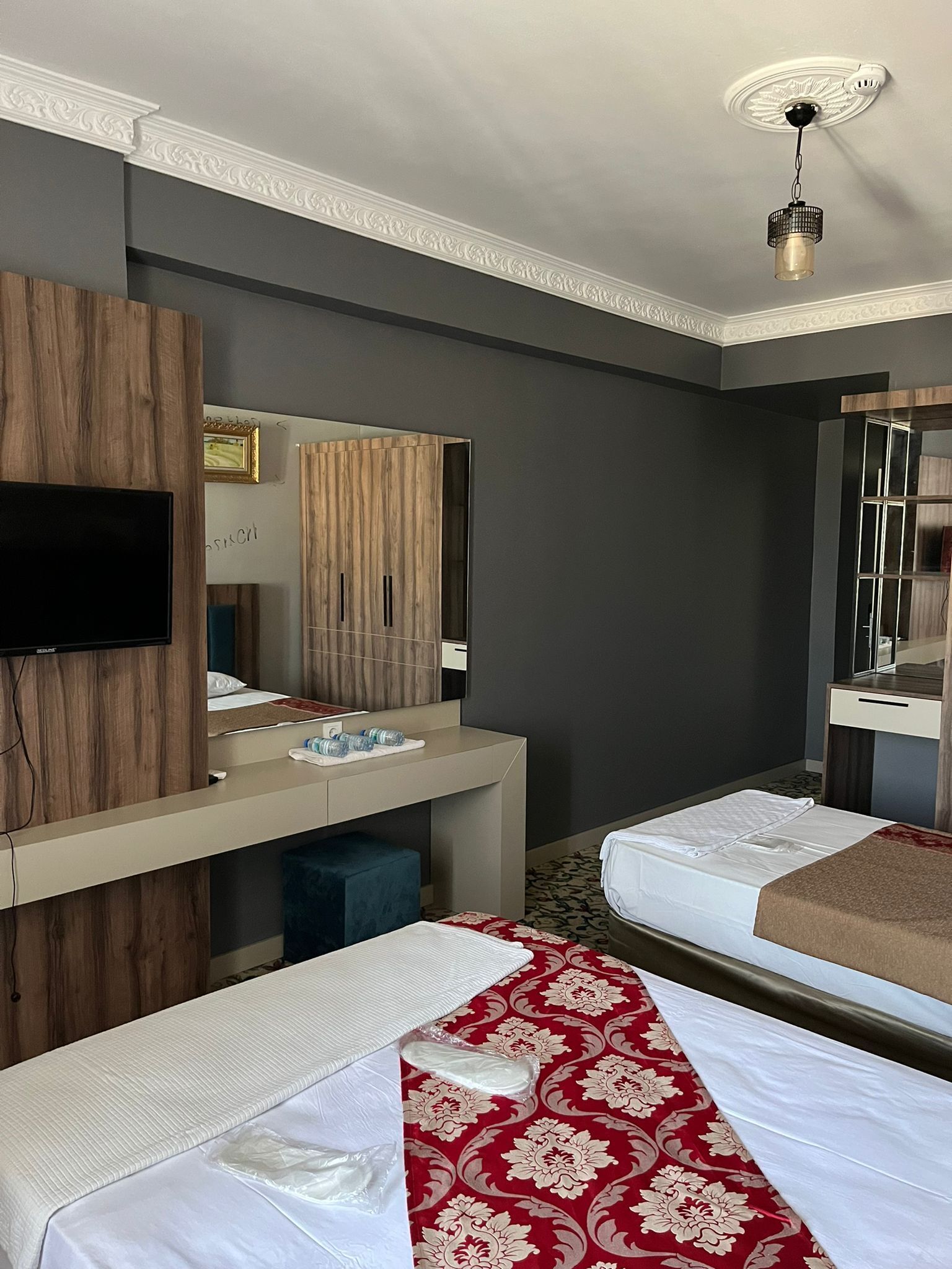 Quarto de hotel com duas camas, uma televisão, um espelho, uma escrivaninha de madeira e um ventilador de teto.