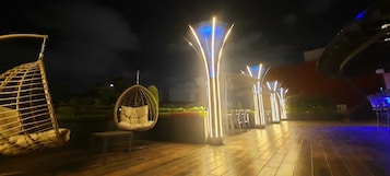 Uma área externa com cadeiras suspensas, colunas iluminadas e um deck de madeira.