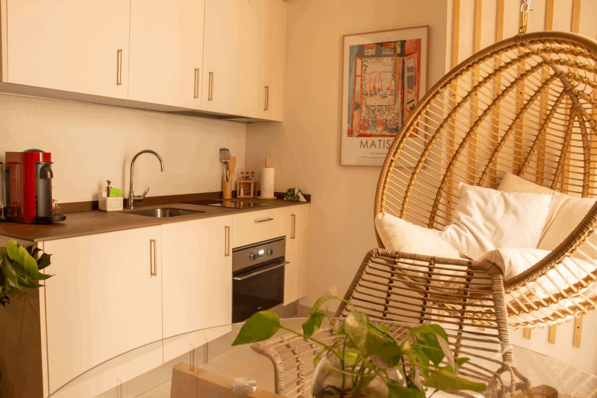 Uma cozinha moderna com armários brancos, forno embutido e uma cadeira de vime suspensa com almofadas.