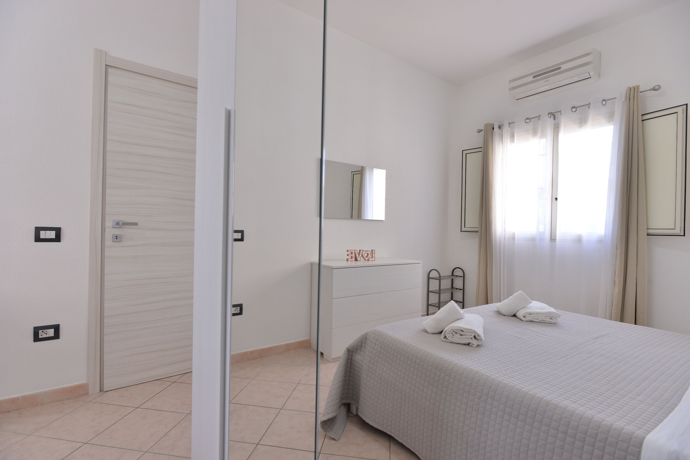 Quarto com cama, mesa de cabeceira, espelho, guarda-roupa e janela com cortinas.
