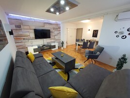 Uma sala de estar moderna com parede de pedra, TV de tela plana, sofá cinza com almofadas amarelas e uma área de jantar ao fundo.