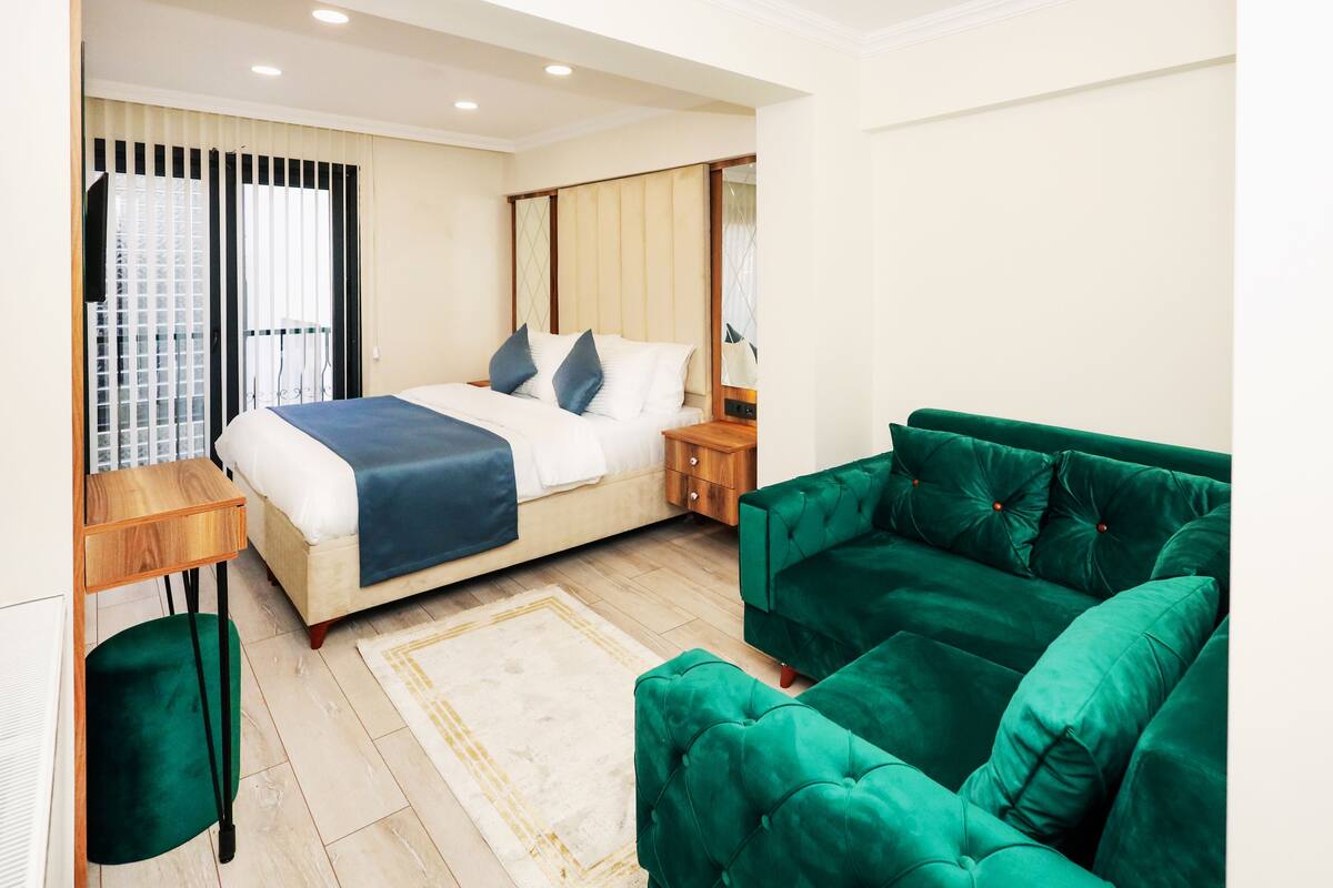 Quarto de hotel moderno com uma cama, um sofá verde, uma mesa de cabeceira de madeira e um espelho grande.