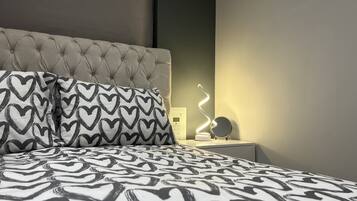 Quarto com cabeceira estofada, almofadas com padrão de coração e mesa de cabeceira com espelho e luminária.