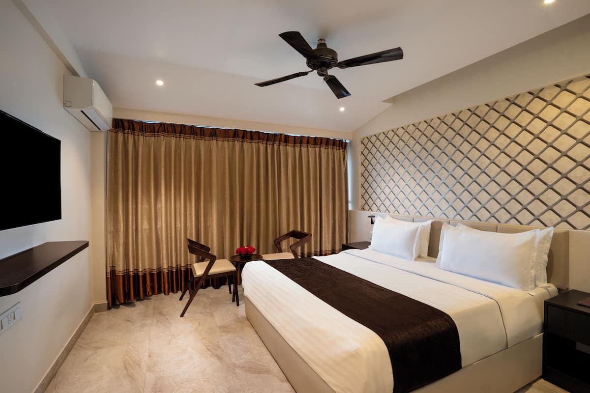 Quarto de hotel com cama, ventilador de teto, televisão, escrivaninha com cadeira e janela com cortinas.