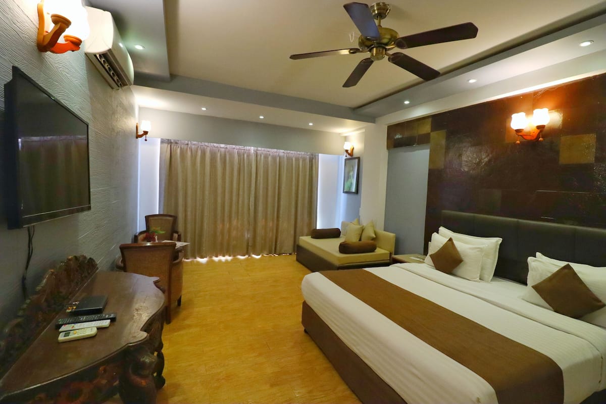 Quarto de hotel com uma cama grande, uma mesa com televisão de tela plana, um ventilador de teto e uma janela com cortinas.