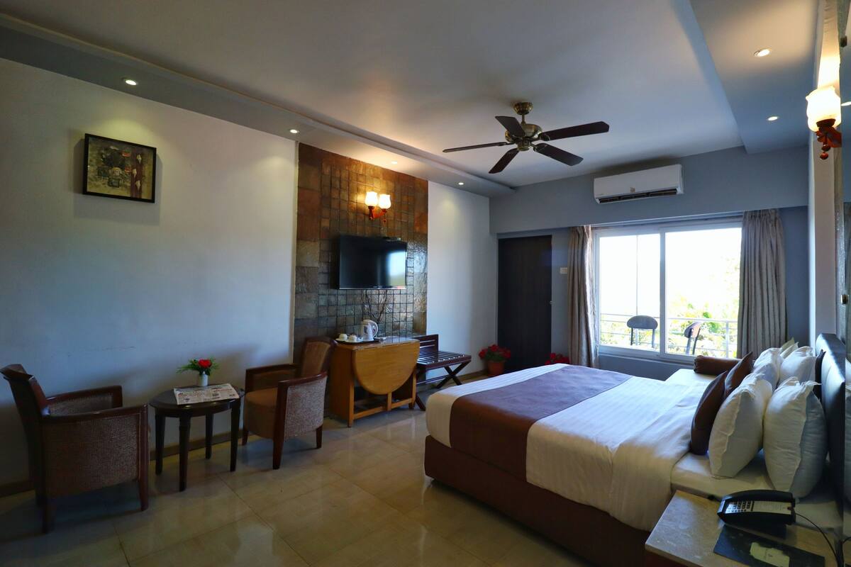 Quarto de hotel com cama, escrivaninha, cadeiras, mesinha com um vaso de flores, ventoinha no teto e uma grande janela com cortinas.