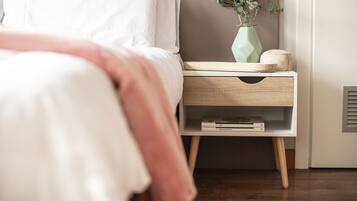 Cama bem arrumada com edredom branco e manta rosa, mesa de cabeceira com um vaso de plantas verdes e mesa de apoio de madeira com livros e uma pequena caixa de madeira.