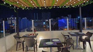 Uma área de refeições ao ar livre com mesas e cadeiras, uma pérgola de madeira com luzes penduradas e vista para a cidade.