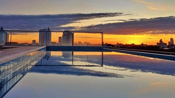 Piscina no terraço com barreira de vidro refletindo o pôr do sol e o horizonte da cidade.