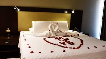 Cama bem arrumada com um arranjo de pétalas de rosa em formato de coração e escultura de toalha sobre um lençol branco.