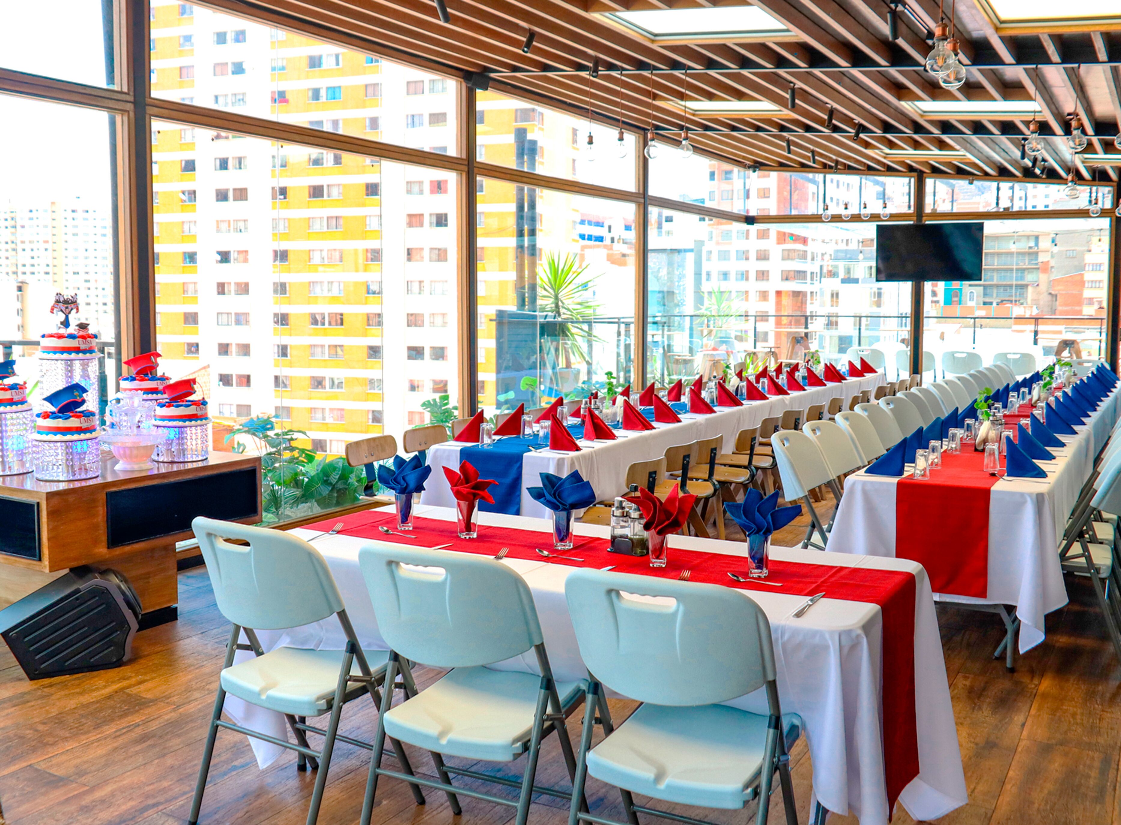 Mesa de jantar comprida montada com cadeiras, guardanapos vermelhos e vista para a cidade através de grandes janelas.