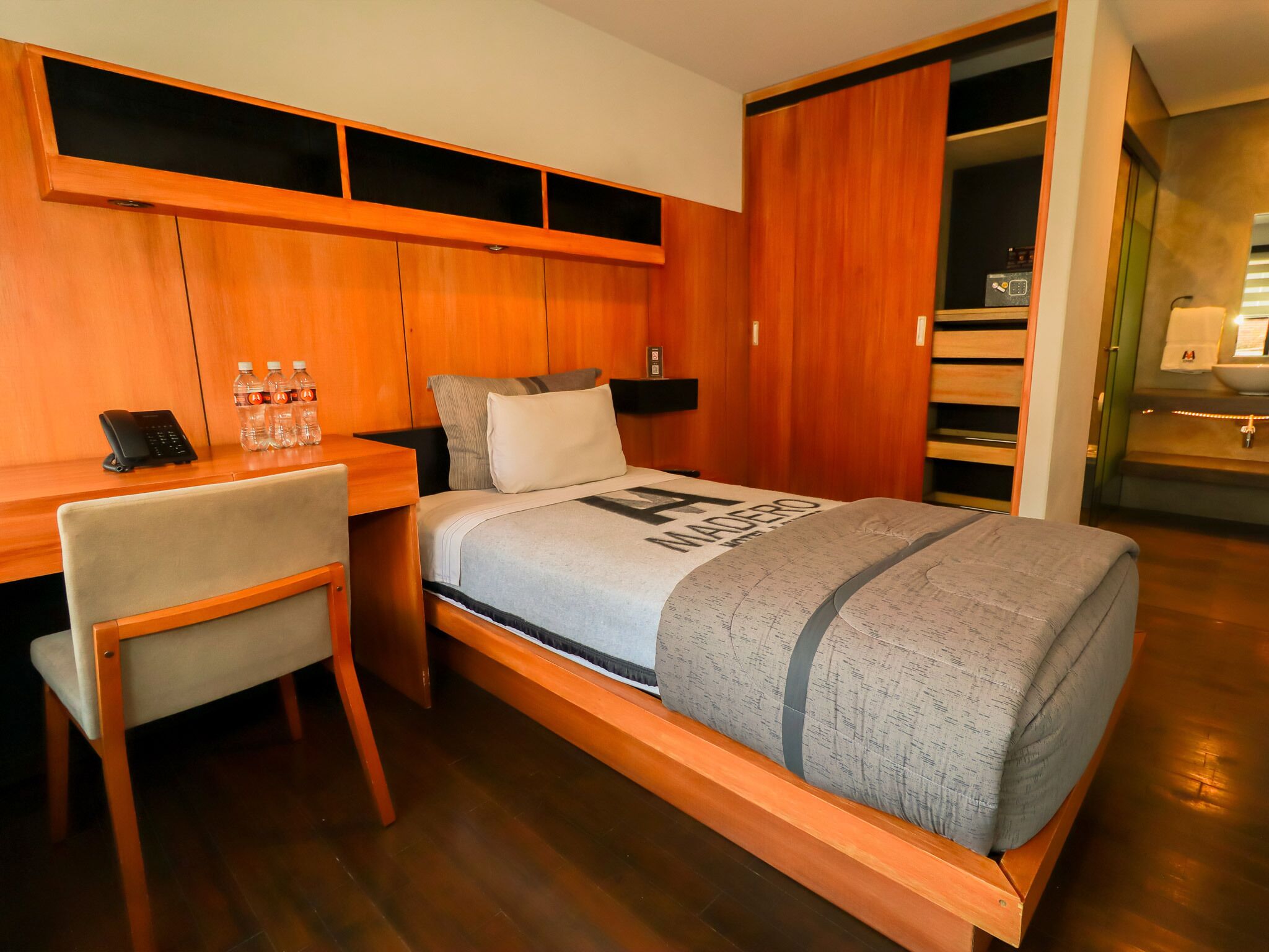 Quarto moderno com cama de madeira, escrivaninha e uma cadeira. É possível ver um minibar e um banheiro ao fundo.