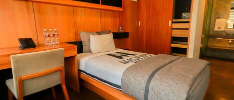 Quarto moderno com cama de madeira, escrivaninha e uma cadeira. É possível ver um minibar e um banheiro ao fundo.
