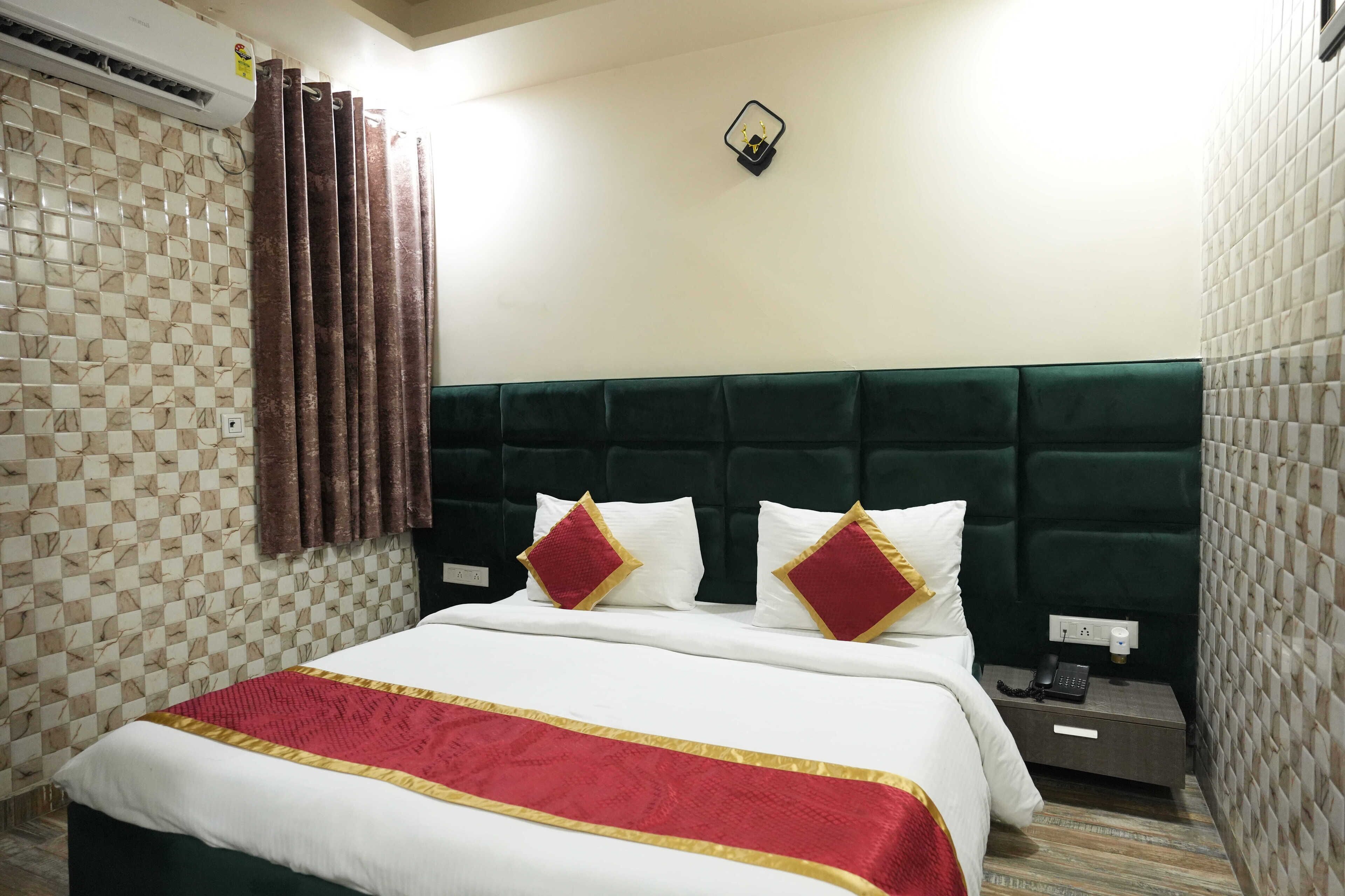 Quarto de hotel com padrão de parede xadrez, cabeceira estofada verde, cama branca com detalhes vermelhos e dourados, e mesa de cabeceira com telefone e copo.