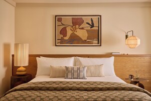Quarto de hotel com cama, almofadas, luminária, uma obra de arte na parede e mesa de cabeceira.
