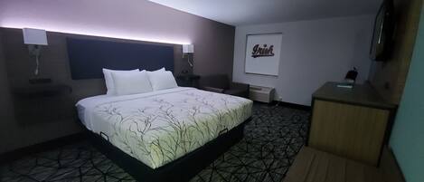 Quarto de hotel com uma cama grande, uma escrivaninha, uma cadeira e uma televisão presa na parede.