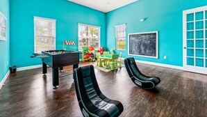 Quarto com paredes verde-água, mesa de futebol de mesa, duas cadeiras pretas, uma mesa com brinquedos infantis e um quadro negro.